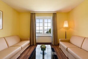 Quarto com dois sofás bege, uma mesa de centro de vidro com uma planta, um abajur e uma janela com cortinas.