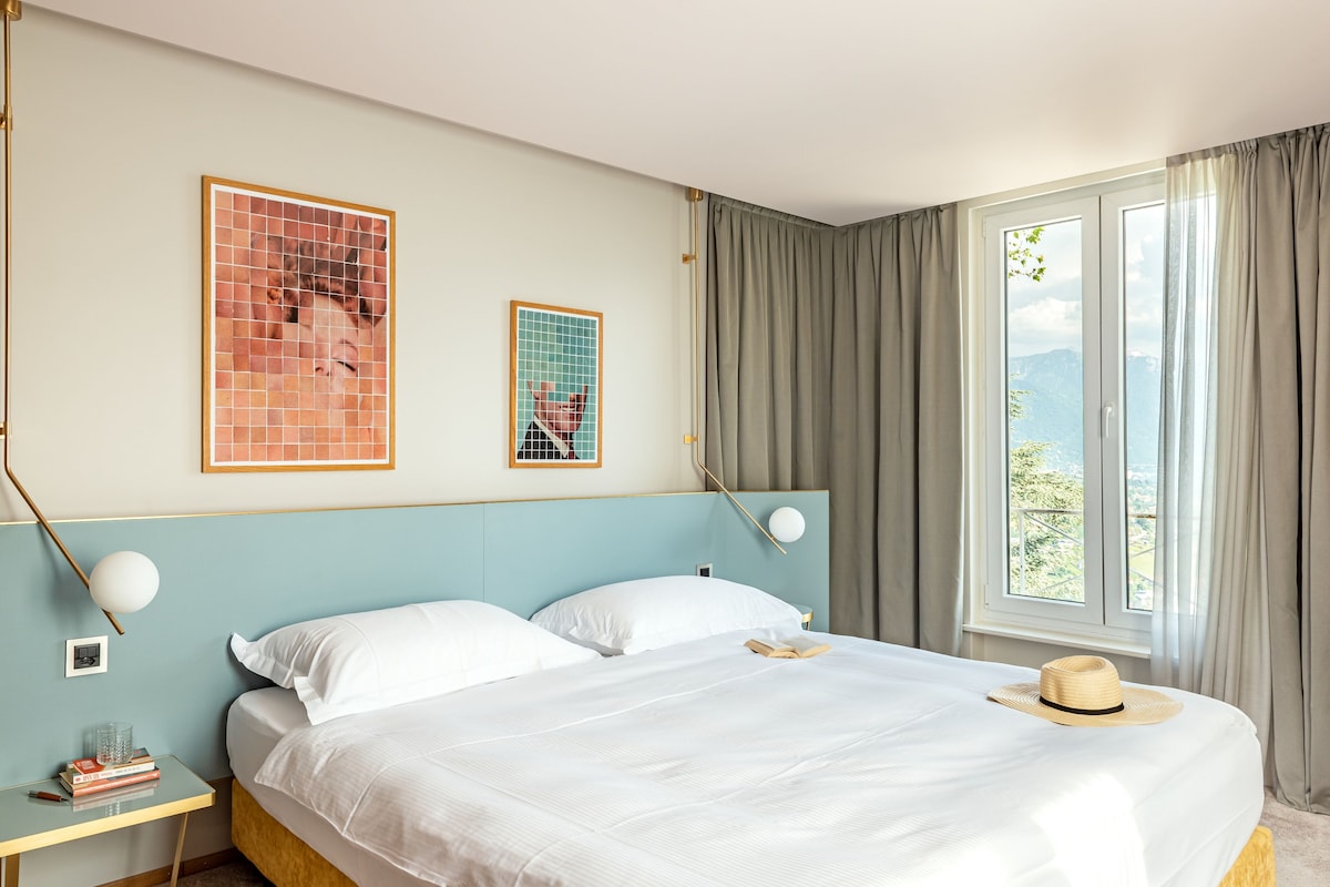 Quarto com cama, mesas de cabeceira, luminária na parede e janela com cortinas.