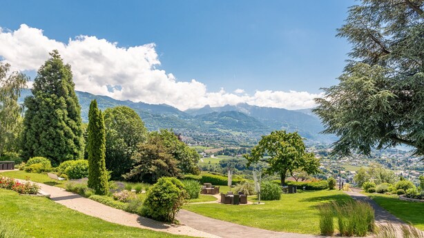 Um jardim com paisagismo, caminhos, áreas de descanso e vista para montanhas ao longe.