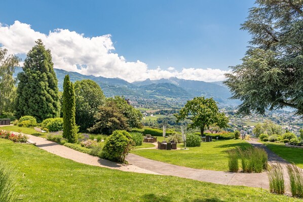 Um jardim com paisagismo, caminhos, áreas de descanso e vista para montanhas ao longe.