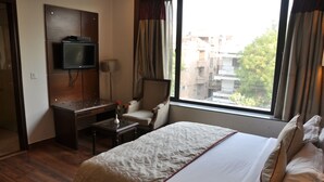 Quarto de hotel com cama, escrivaninha, cadeira, televisão e janela com vista para edifícios.