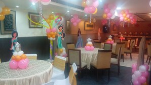 Uma sala de banquetes com mesas redondas cobertas por toalhas brancas, cadeiras com fitas amarelas e balões rosa e brancos.