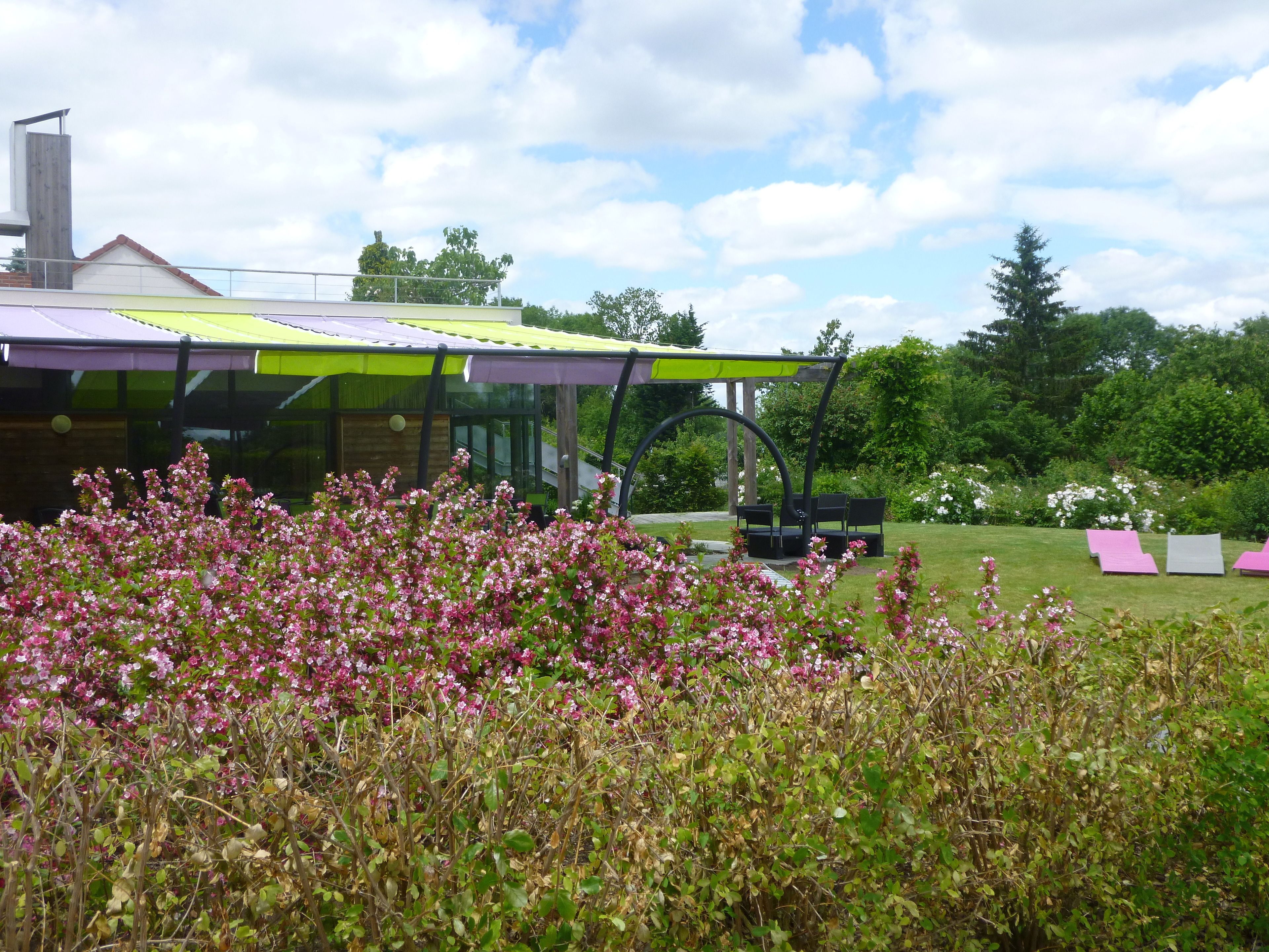Um edifício moderno com fachada de vidro e telhado verde, rodeado por vegetação exuberante e flores cor-de-rosa.