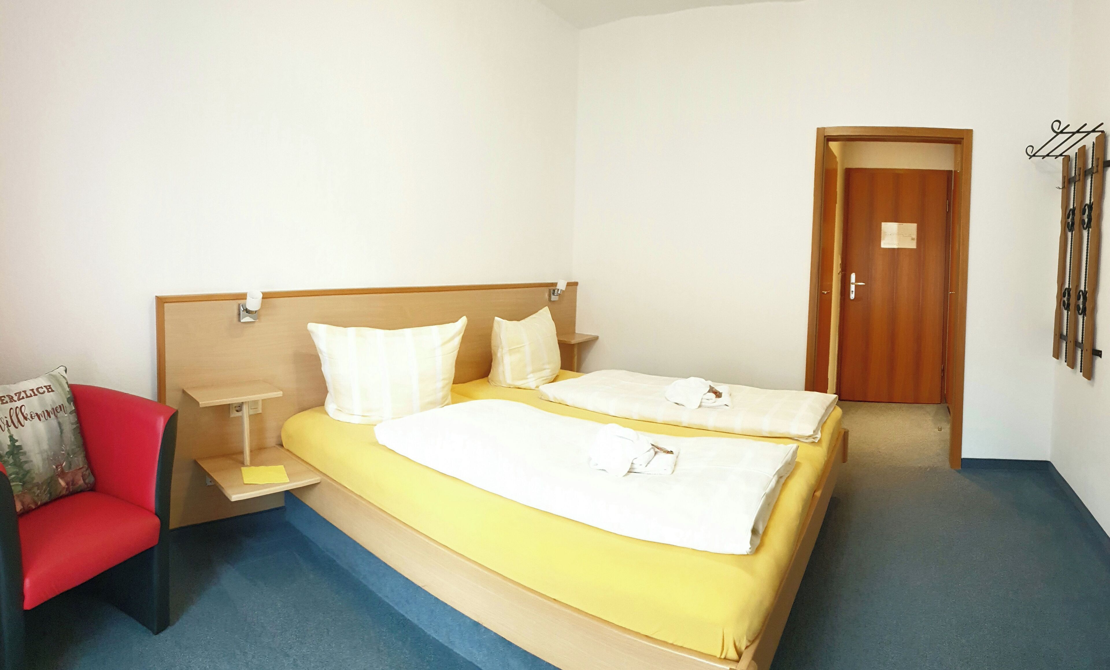 Quarto de hotel com cama de casal, cabeceira de madeira, mesa de cabeceira pequena, uma cadeira vermelha e uma porta com um aviso.