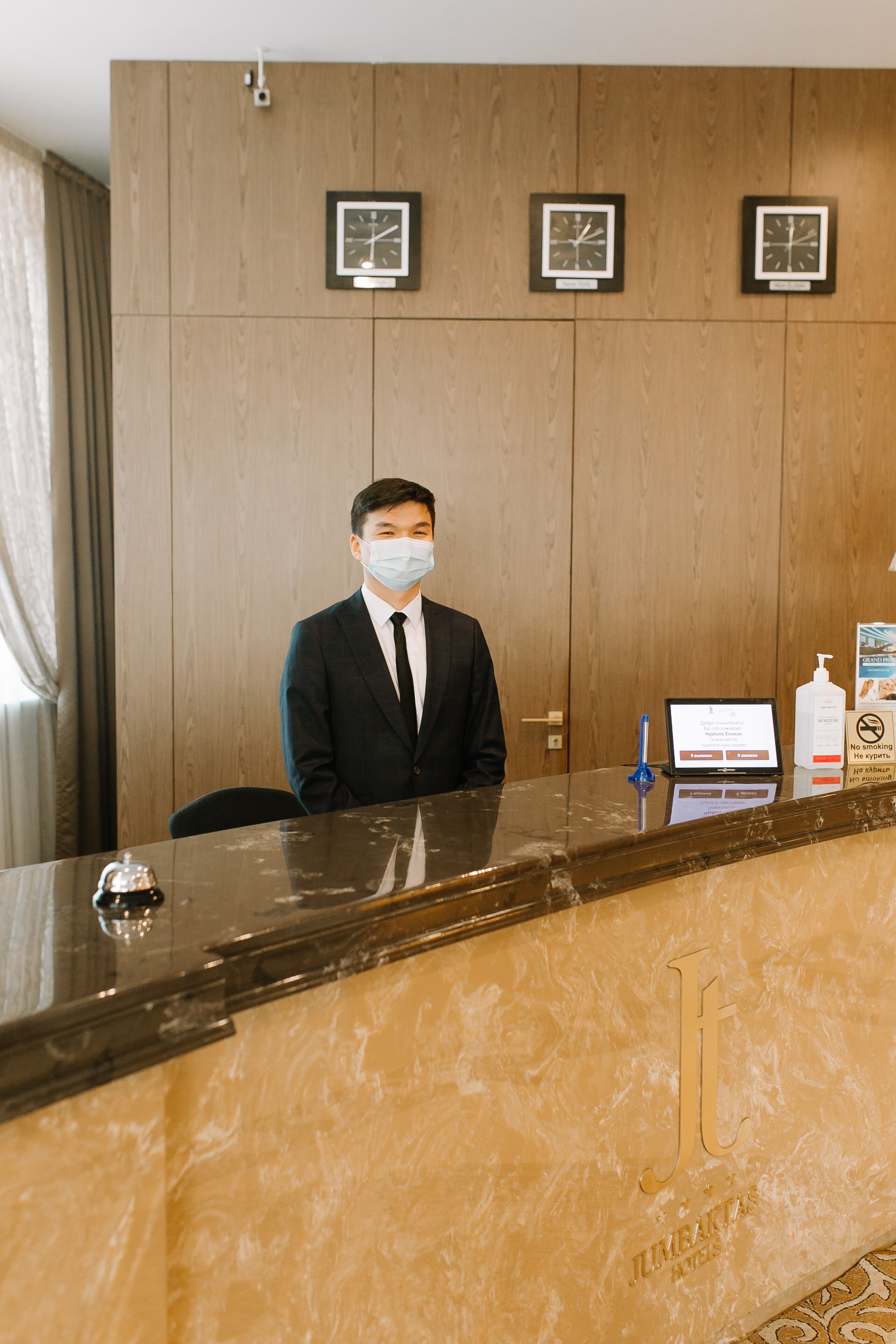 Balcão de recepção de hotel com um homem de terno, painéis de madeira, relógios e um laptop.