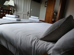 Cama bem arrumada com lençóis brancos e almofada cinza, uma cadeira de madeira com toalha branca, e um abajur sobre uma mesa de cabeceira.