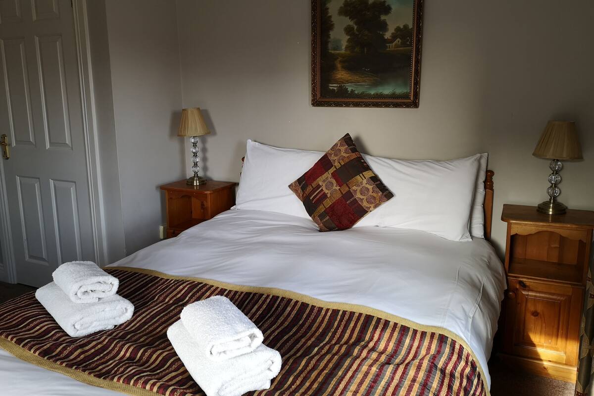 Cama bem arrumada com lençóis brancos, manta decorativa e duas toalhas enroladas. Há duas mesas de cabeceira com abajures, um quadro na parede e uma porta à esquerda.
