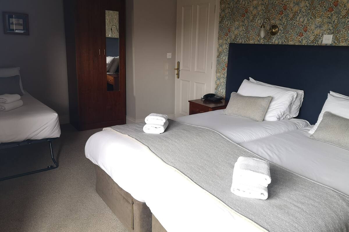 Quarto de hotel com duas camas, um guarda-roupa de madeira, uma mesa de cabeceira com telefone e papel de parede estampado.