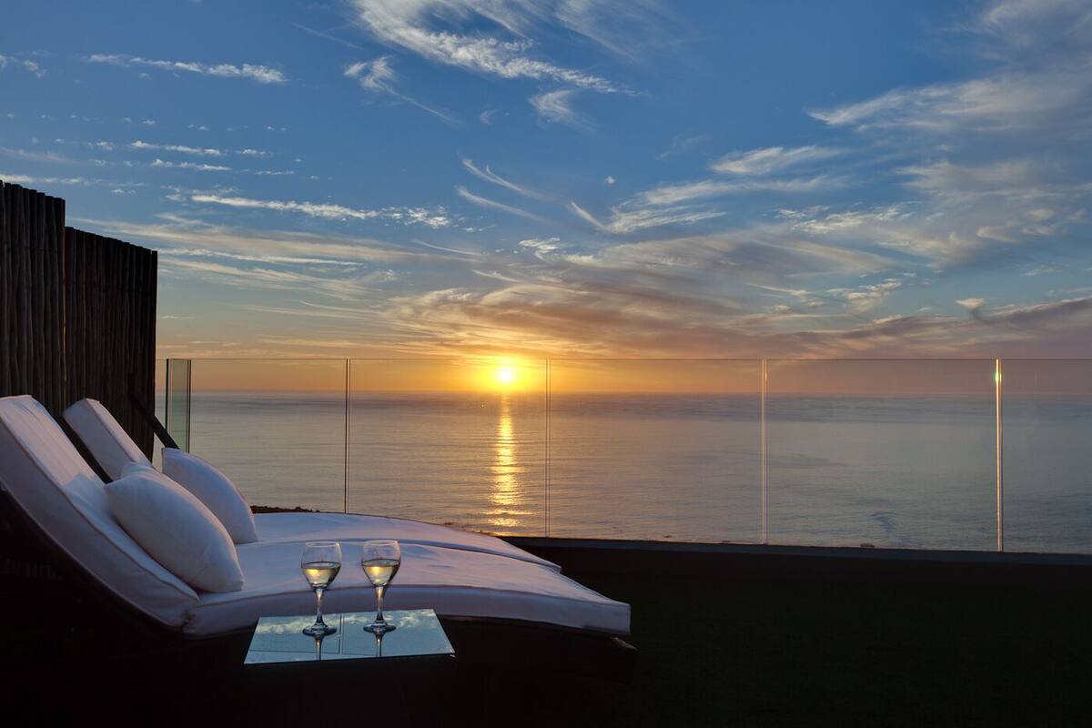 Vista do pôr do sol a partir de uma varanda com cadeiras de descanso e duas taças de vinho.