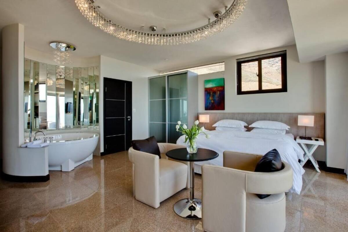 Quarto de hotel moderno com uma cama grande, um espaço de estar com mesa, uma banheira e um espelho amplo.