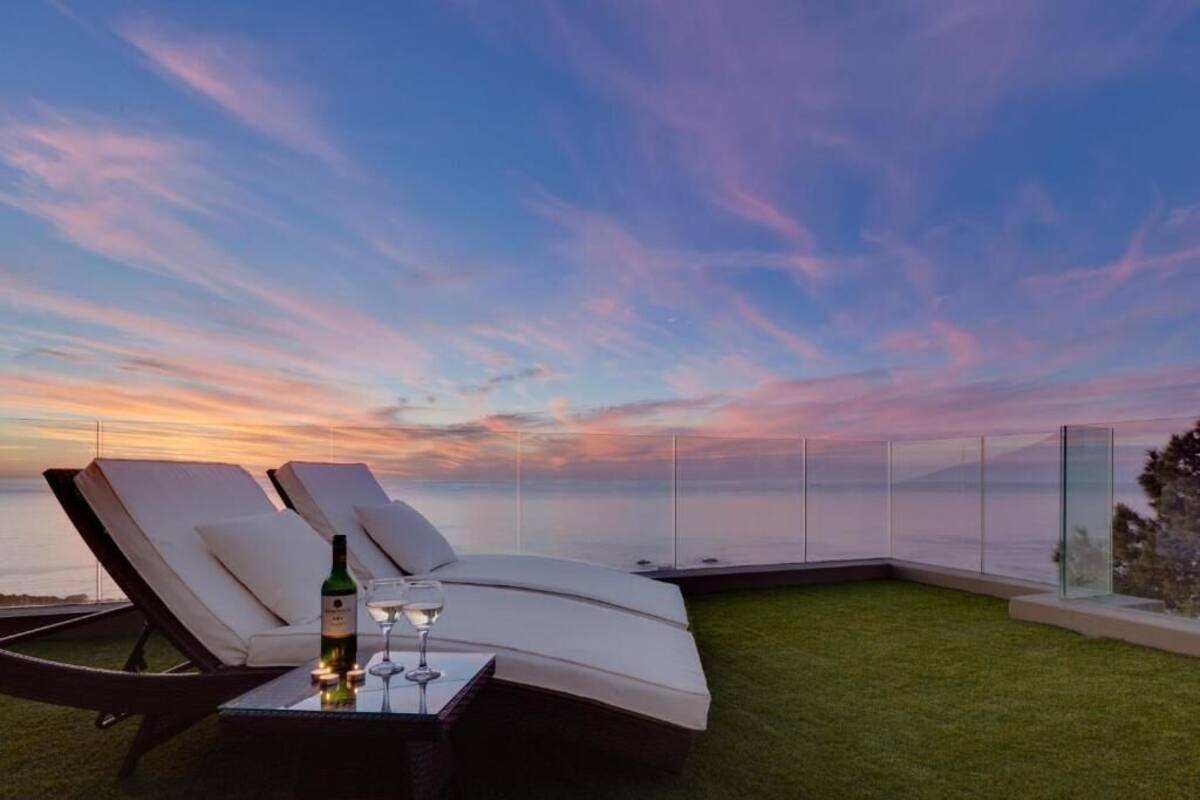 Vista do pôr do sol vista de uma varanda, com uma mesa, duas poltronas de descanso e uma garrafa de vinho.