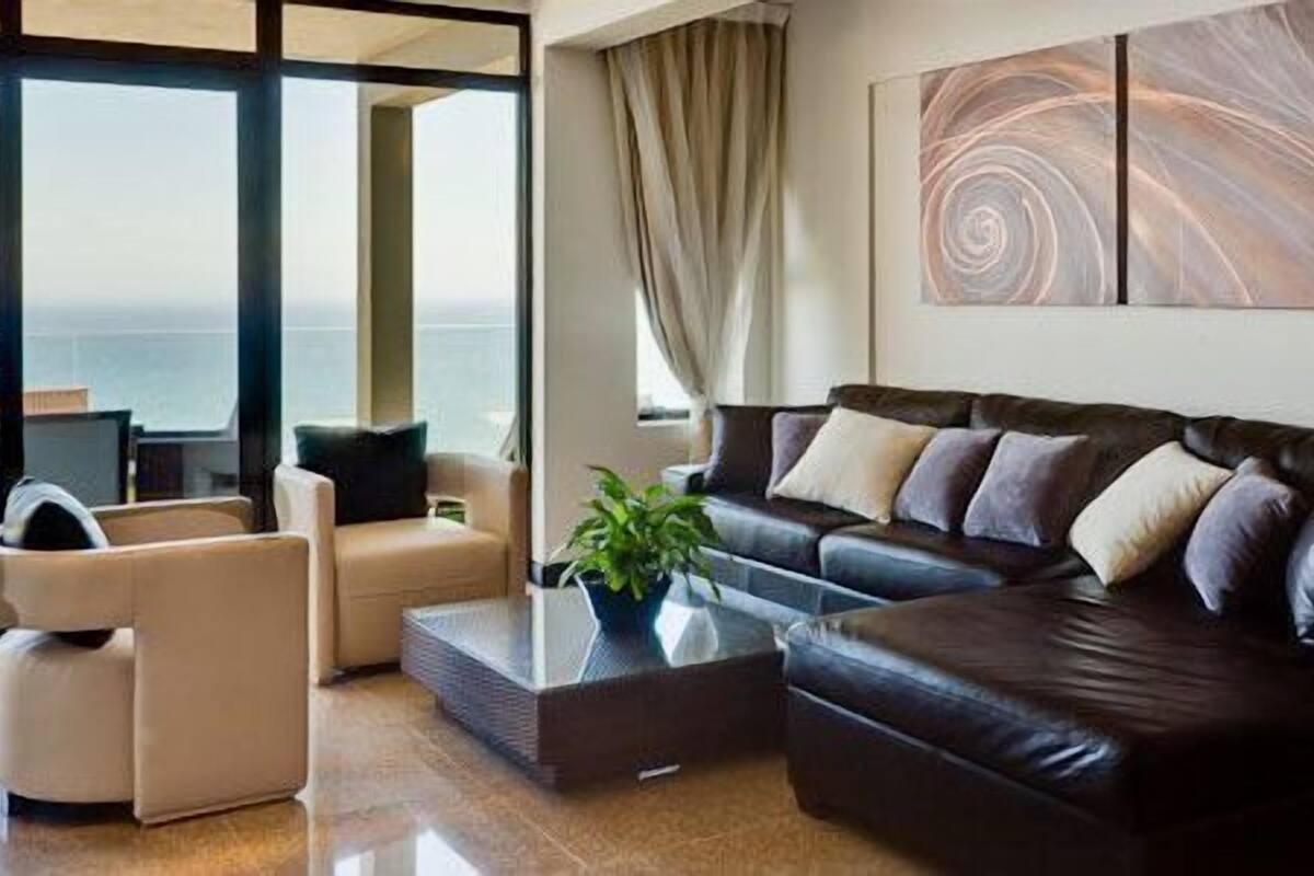 Uma sala de estar moderna com um sofá de couro preto, poltronas brancas e um vidro que dá para uma varanda com vista para o mar.