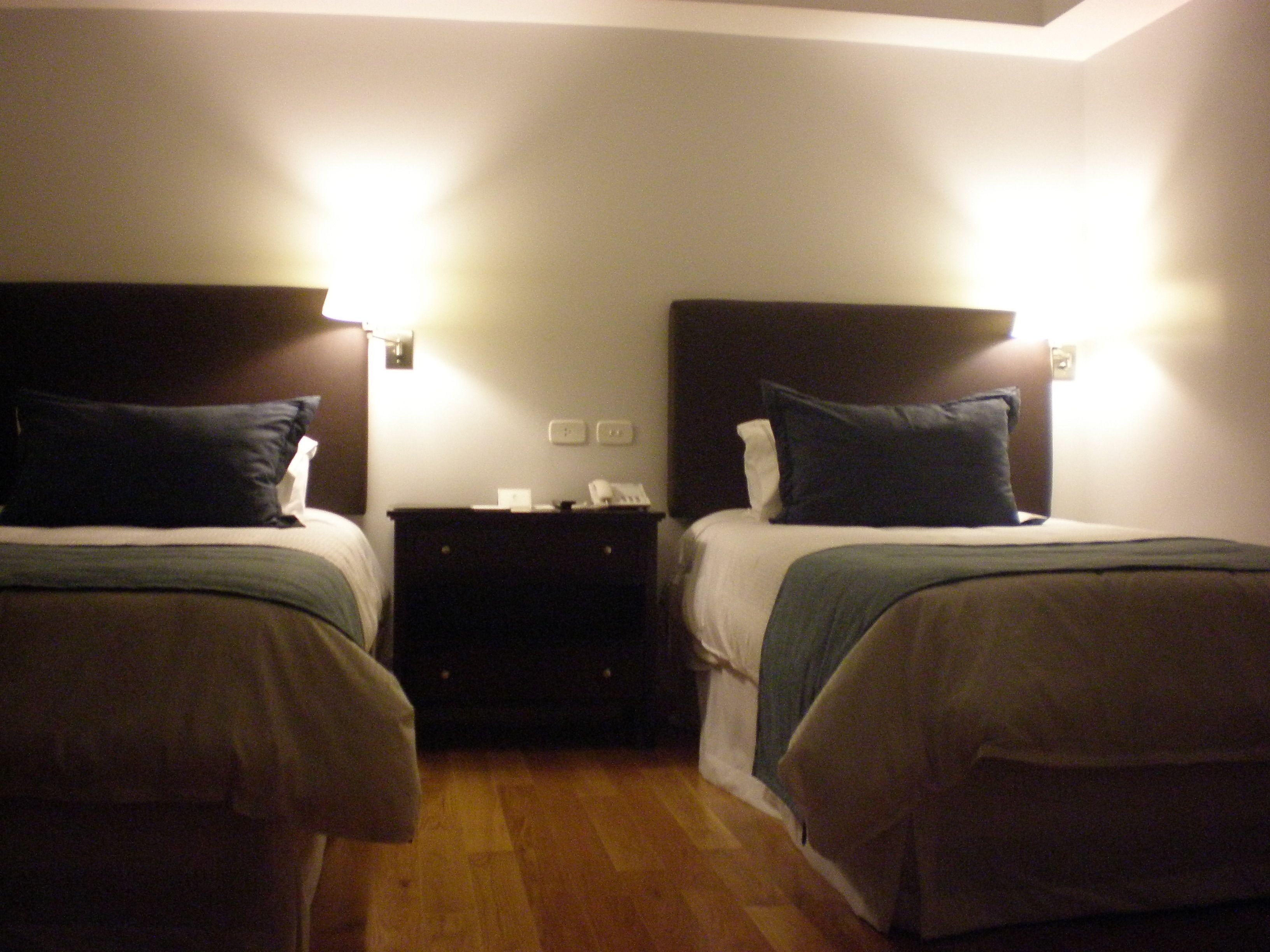 Quarto duplo com duas camas, cada uma com travesseiro e cobertor. Há uma mesinha de cabeceira entre as camas com uma lâmpada e um telefone.