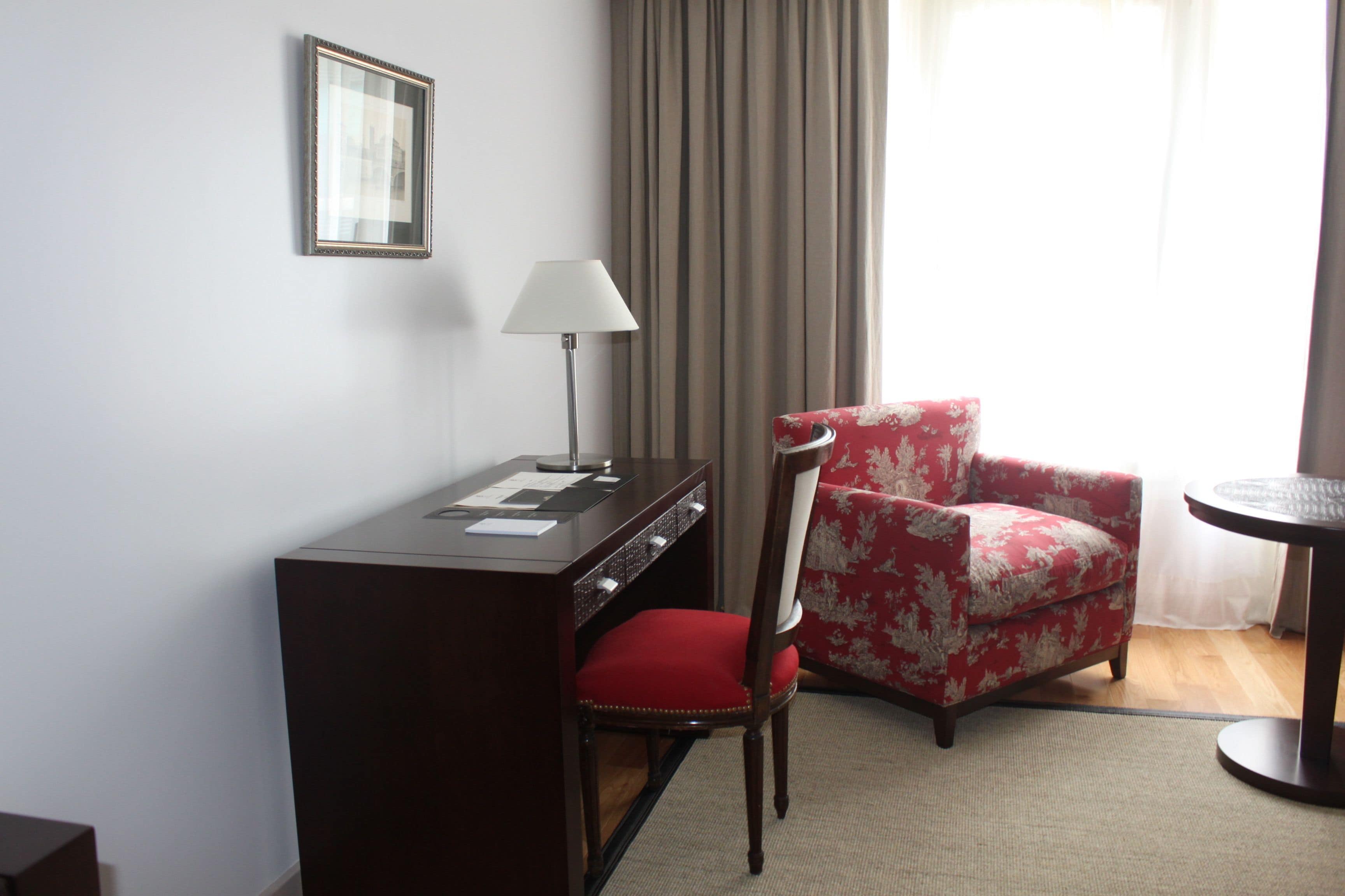 Quarto de hotel com escrivaninha, cadeira, luminária e uma mesa redonda.