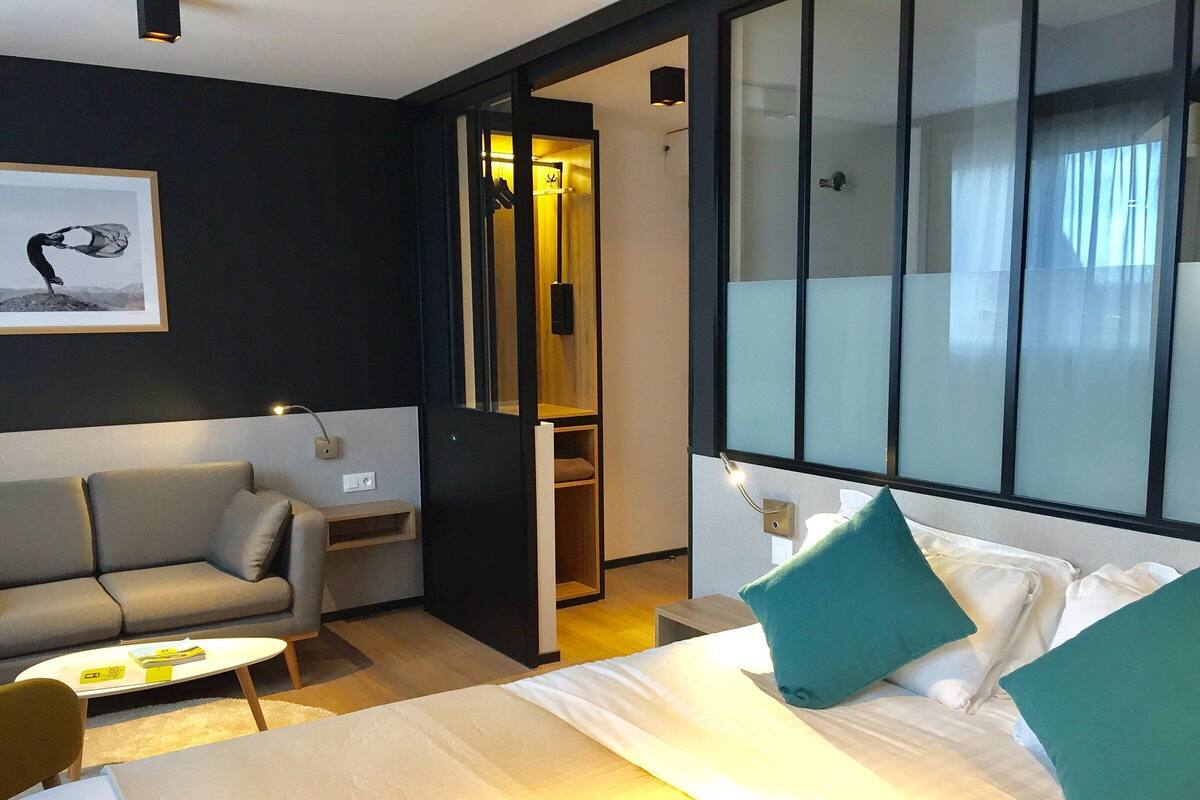Quarto de hotel moderno com cama, sofá, uma mesinha e espelho.