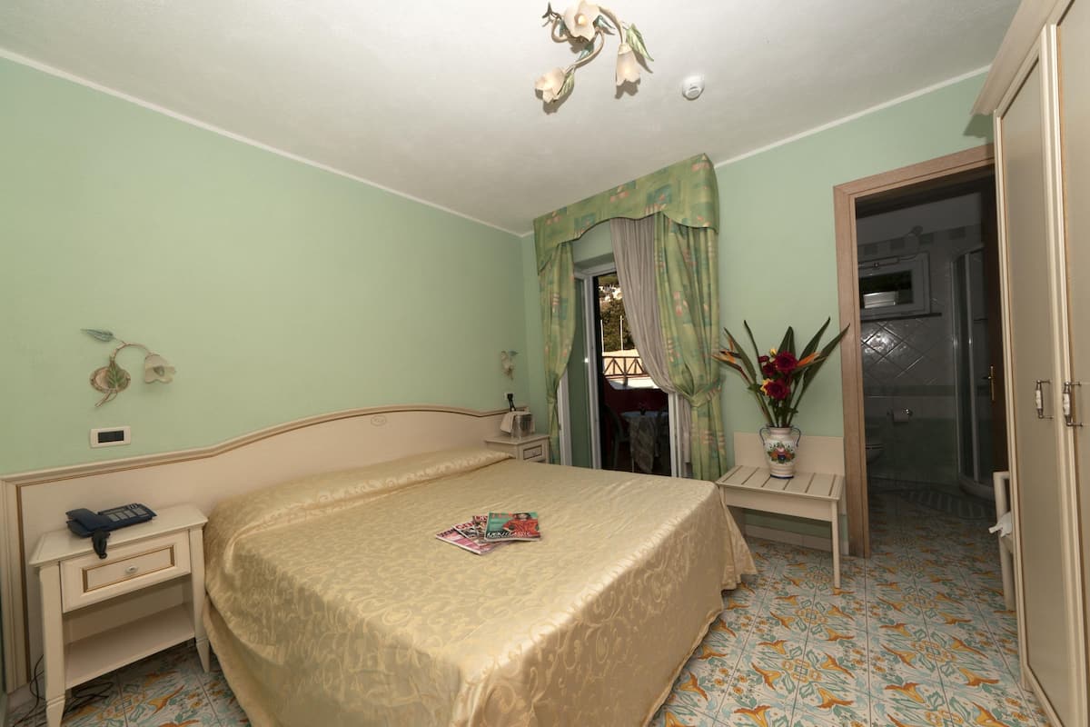 Quarto com cama, mesas de cabeceira, escrivaninha, cadeira, janela com cortinas, porta e banheiro visível através da porta.