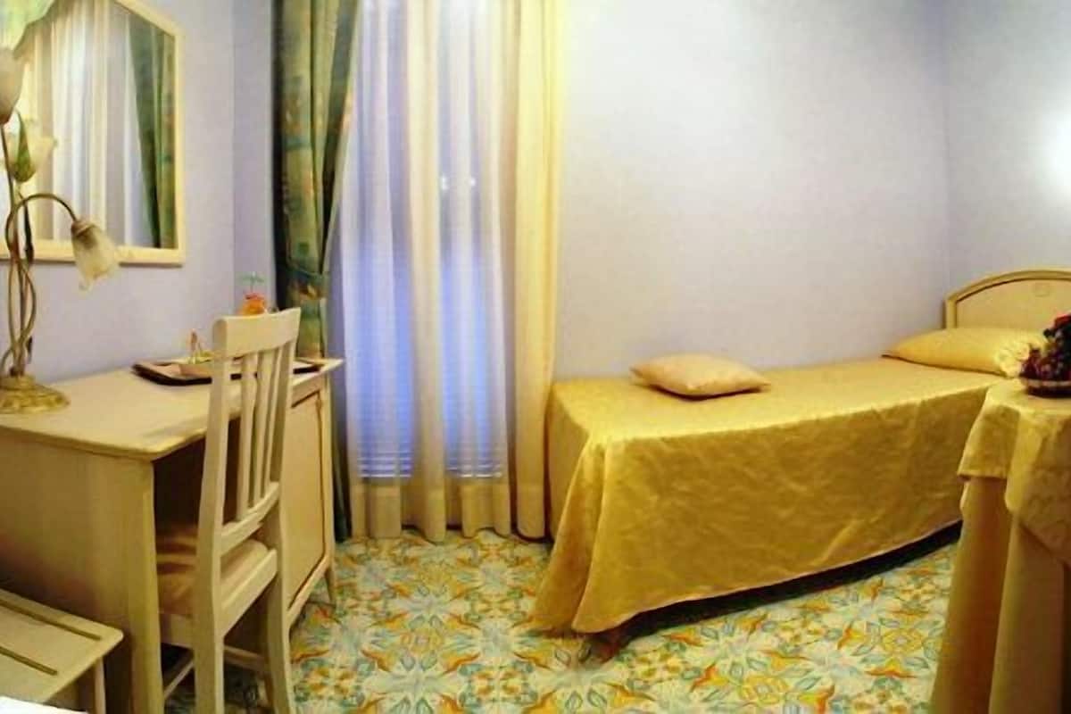 Quarto pequeno e bem arrumado com uma cama de solteiro, uma escrivaninha e uma cadeira.