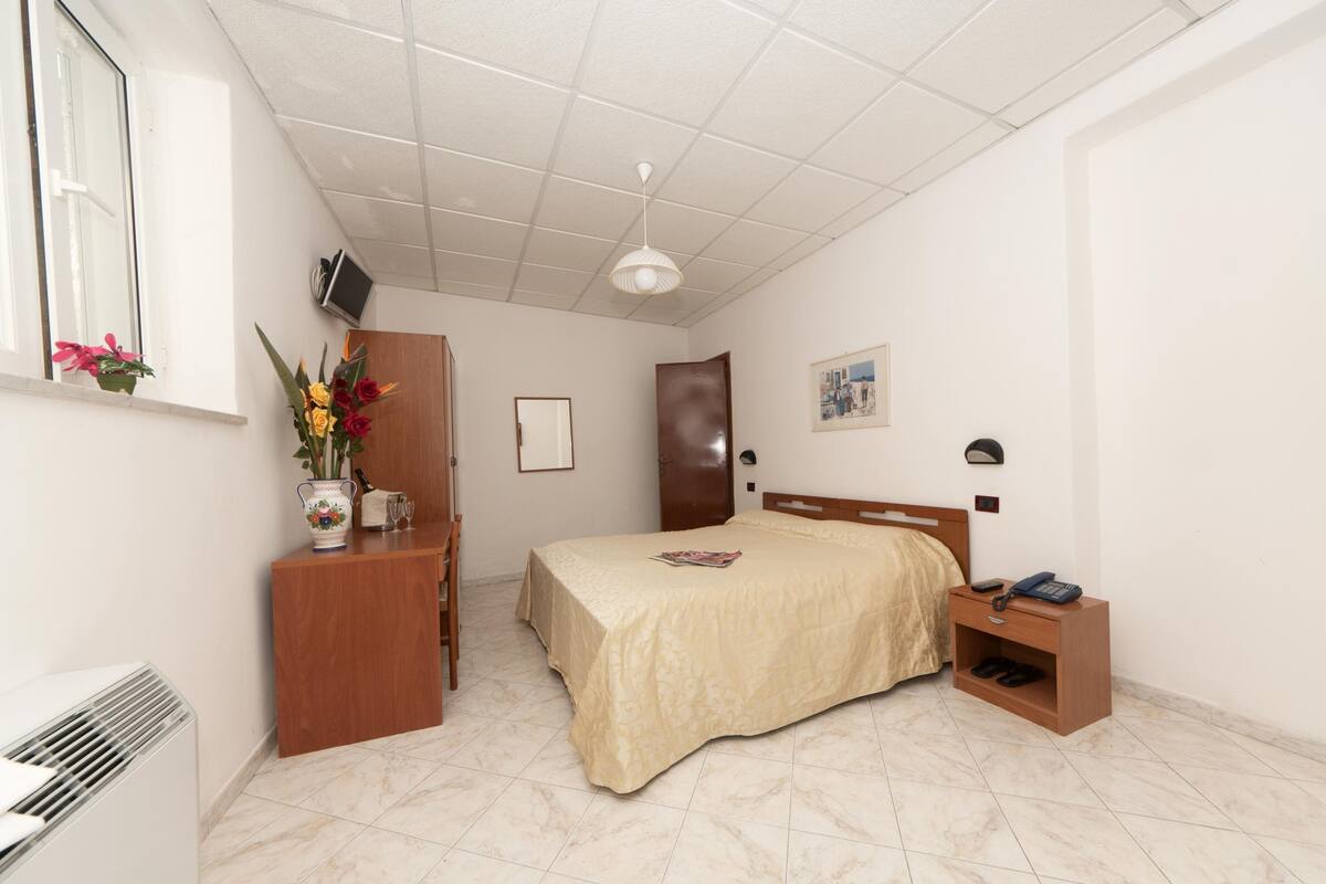 Quarto com cama, mesa de cabeceira, guarda-roupa e pequena escrivaninha com cadeira.