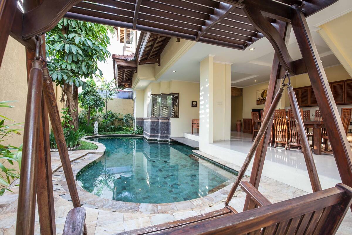 Uma área de piscina com deck de ladrilhos de pedra, cadeiras de madeira e uma estrutura de pérgola.