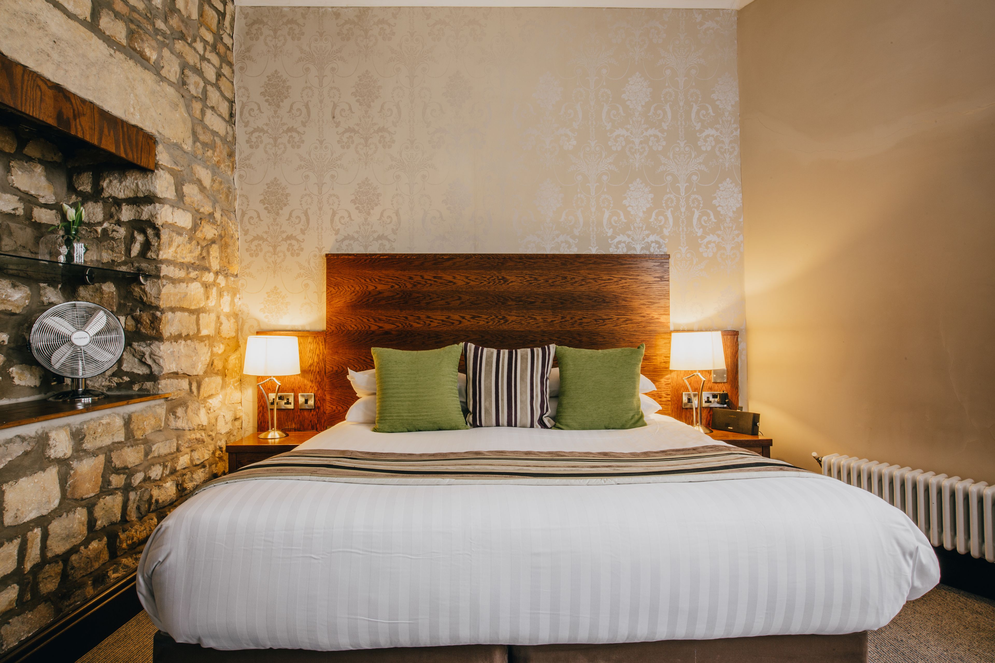 Quarto de hotel com uma cama grande, duas luminárias de cabeceira, um relógio e uma parede de pedra.