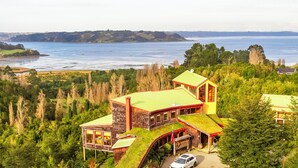 Uma casa com telhado verde, rodeada por uma floresta densa, perto de um corpo d'água.