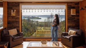 Quarto com uma janela ampla que oferece uma vista panorâmica de um lago e colinas. Há dois sofás de couro e uma mesa de centro de vidro no quarto.