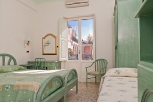 Quarto com duas camas, uma escrivaninha e uma cadeira. Há uma janela com vista para prédios.