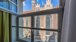 Uma janela com grade de metal, vista de um edifício histórico de tijolos com torres pontiagudas e uma cruz no topo, e um céu azul límpido.