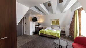 Quarto de hotel moderno com teto alto, cama com colcha verde, escrivaninha com luminária, ventilador e uma cadeira vermelha.