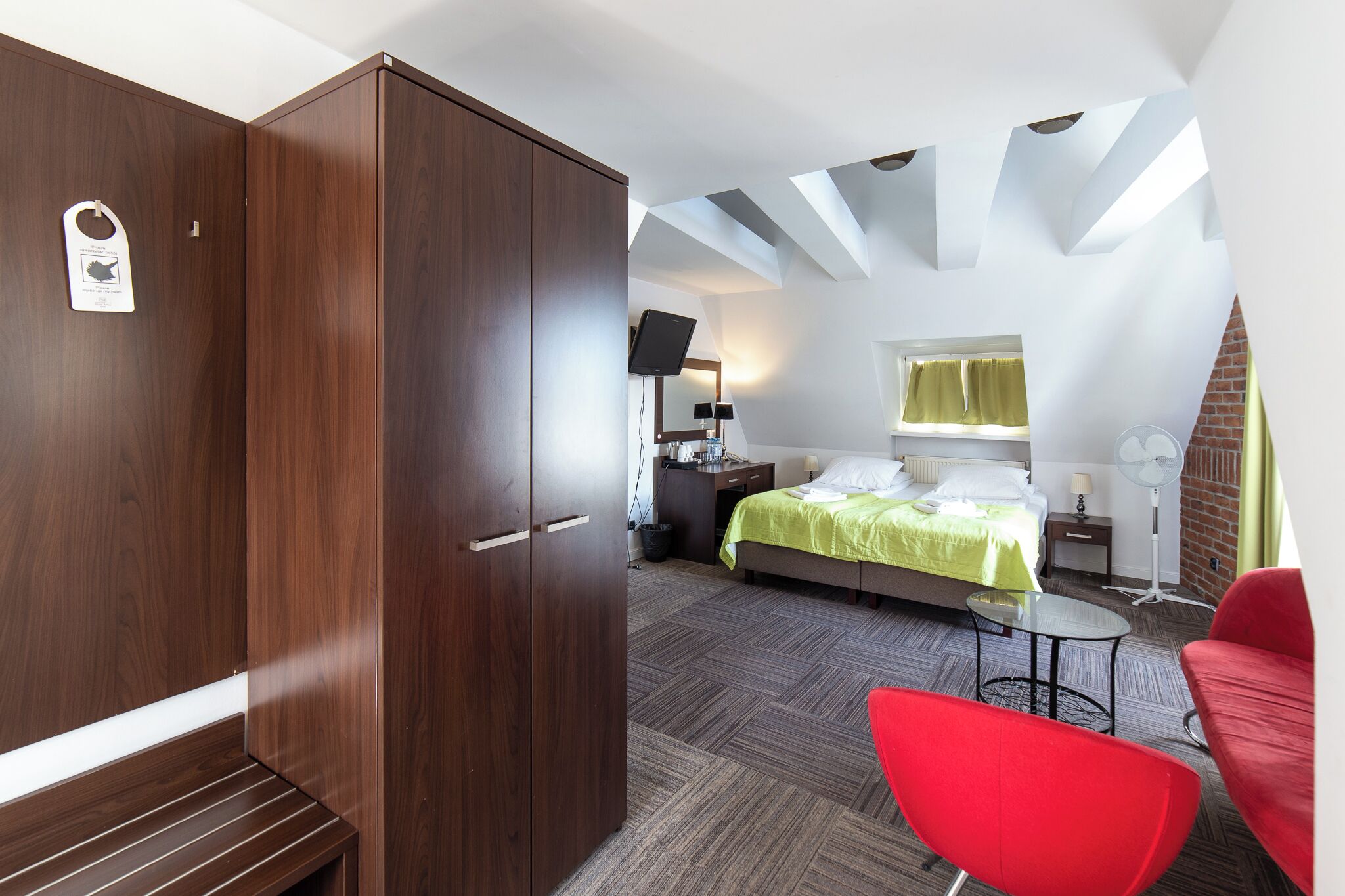 Quarto de hotel com um guarda-roupa espaçoso, uma cama com colcha verde, uma cadeira vermelha, uma mesa pequena e um ventilador de teto.