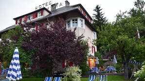 Um hotel com área de jardim, apresentando cadeiras e guarda-sóis de lounge listrados em azul e branco, uma grande árvore com folhas roxas e uma escadaria de pedra que leva à entrada.