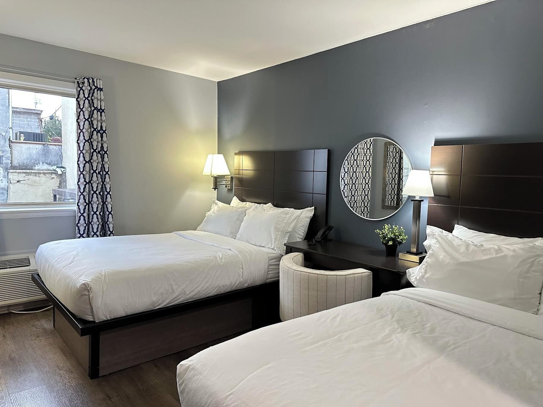 Quarto de hotel com duas camas, uma mesa de cabeceira com luminária, espelho fixado na parede e janela com cortinas.