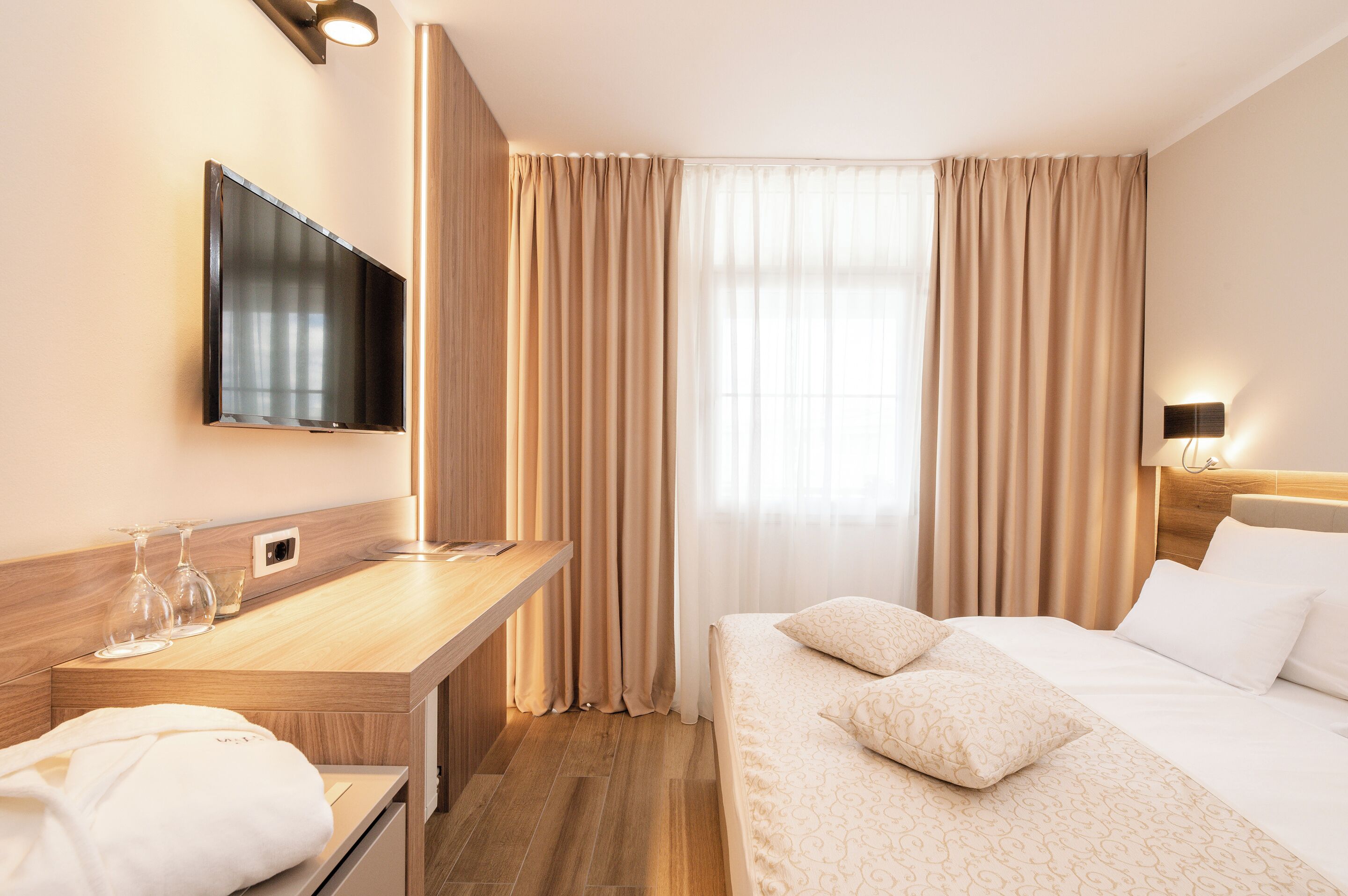 Quarto de hotel com cama, escrivaninha, televisão e janela com cortinas.