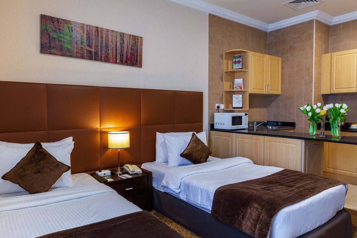Quarto de hotel com duas camas, uma mesa de cabeceira com abajur, micro-ondas e pequena cozinha com armários de madeira e bancada.