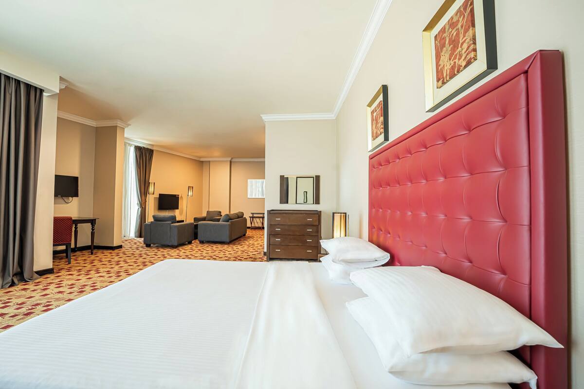 Quarto de hotel com uma cama grande, cabeceira vermelha, escrivaninha com cadeira, sofá e cómoda.