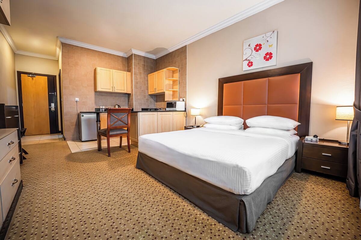 Quarto de hotel com cama, escrivaninha, cadeira, cozinha compacta e banheiro.