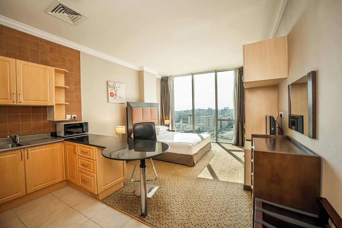 Quarto de hotel com cozinha compacta, cama, janela grande com vista para a cidade e televisão de tela plana.