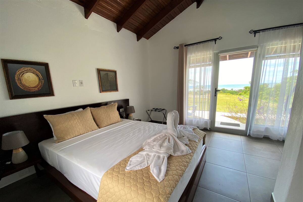 Quarto de hotel com uma cama grande, cabeceira, mesa de cabeceira com abajur e vista para o exterior através de uma porta de correr de vidro.