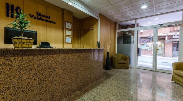 Uma área de recepção de hotel com balcão de mármore, uma planta, certificados emoldurados e uma cadeira.