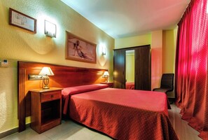 Quarto de hotel com cama, duas luminárias de cabeceira, mesa de cabeceira, uma cadeira, porta e um quadro na parede.