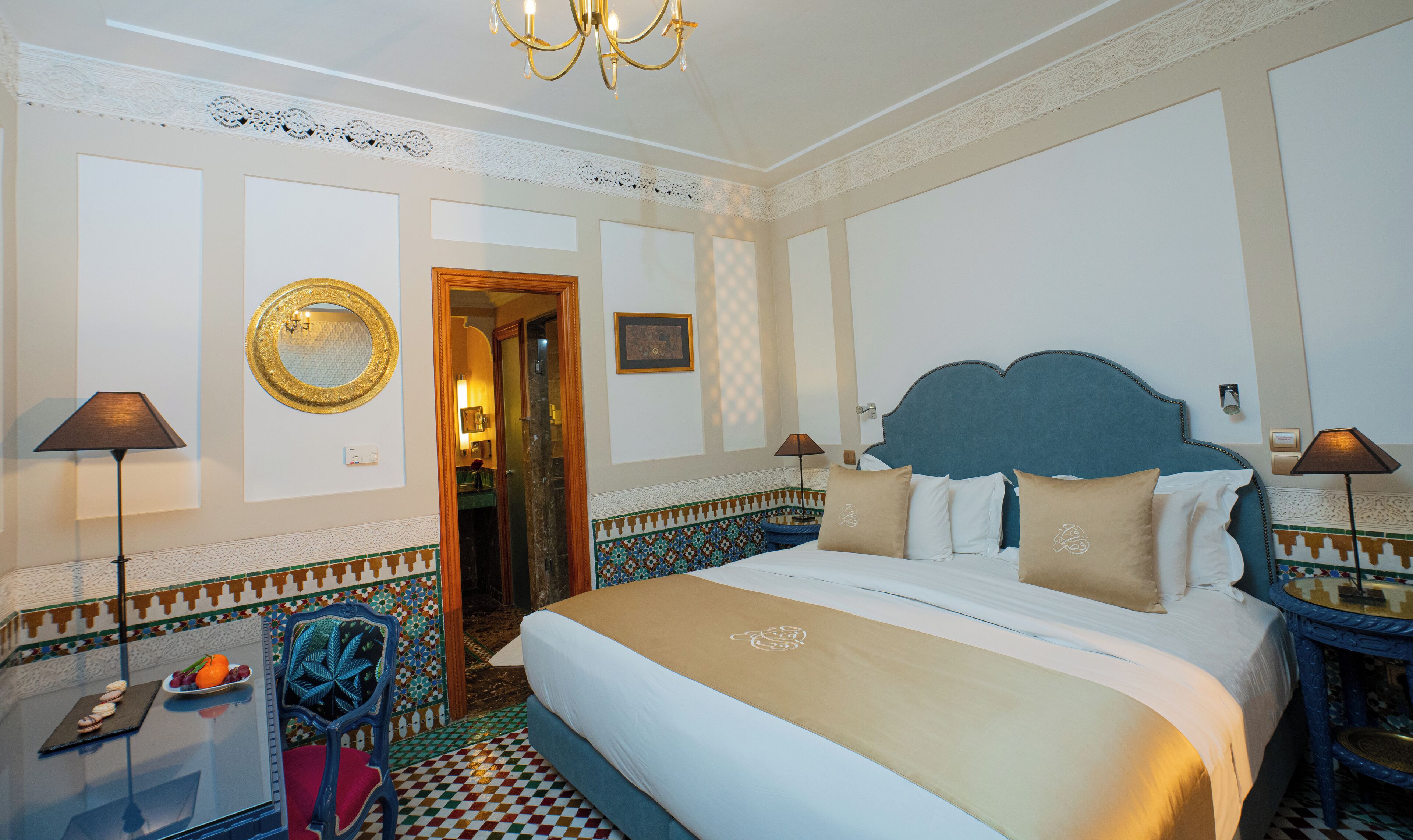 Quarto de hotel com uma cama grande, duas mesas de cabeceira com luminárias, uma cadeira, um espelho e piso estampado.