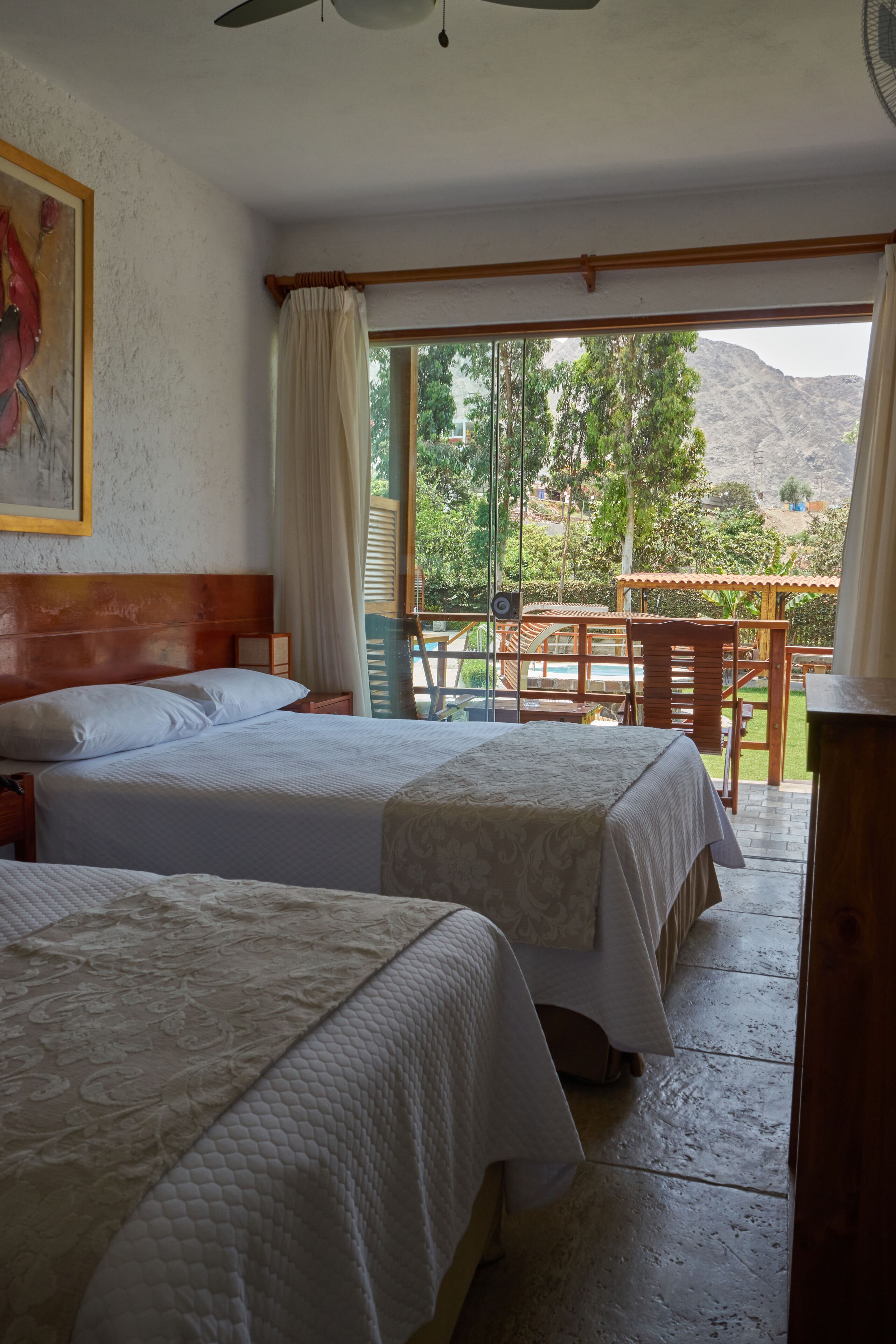 Quarto com duas camas, cabeceira de madeira, quadro na parede e varanda com vista para árvores e montanhas.