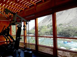 Uma academia com equipamentos de ginástica e amplas janelas com vista para um rio e montanhas.