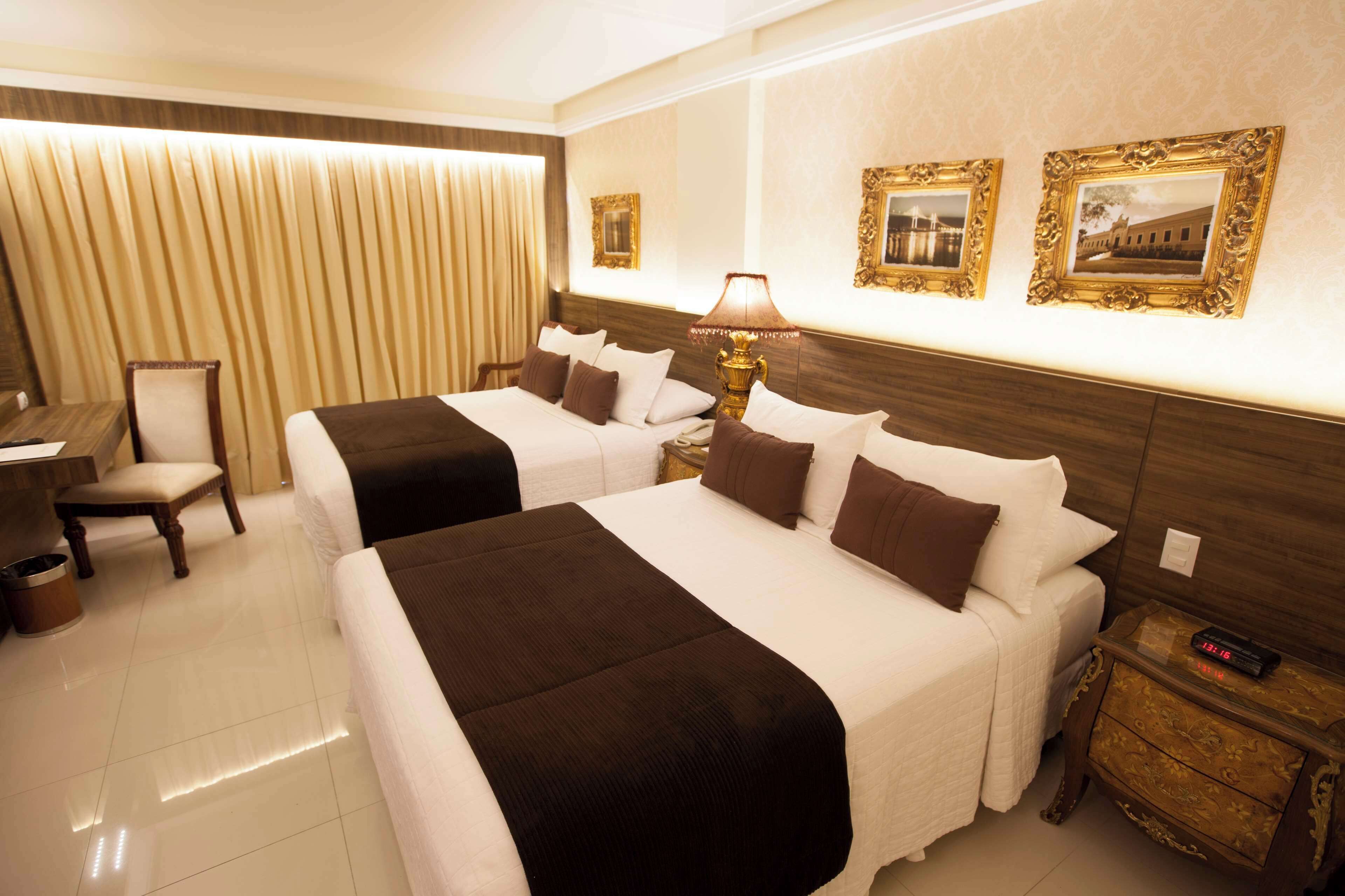 Quarto de hotel com duas camas, uma escrivaninha, uma cadeira e uma mesa de cabeceira com relógio.
