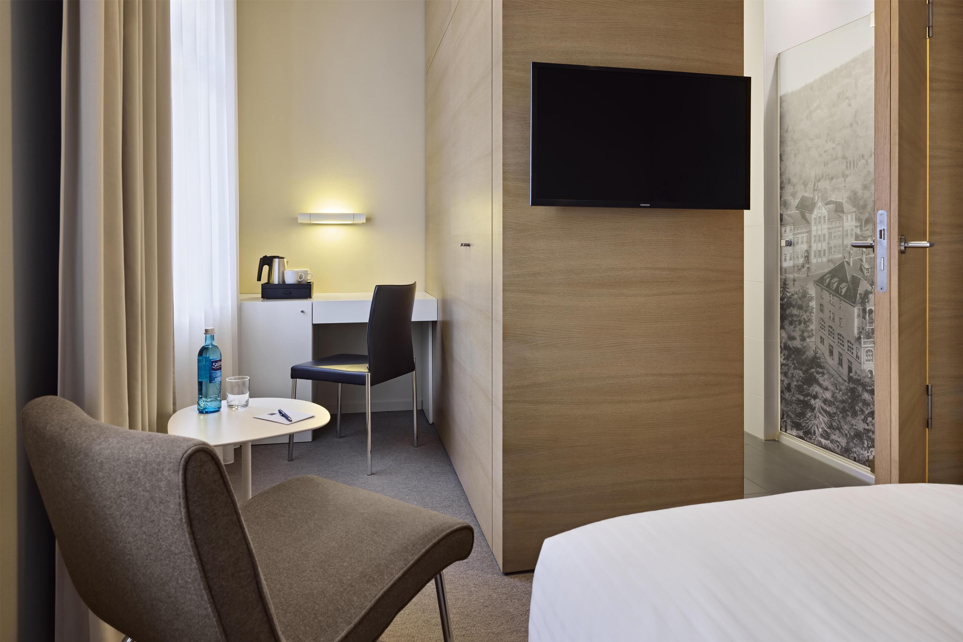 Quarto de hotel com cama, escrivaninha com cadeira, televisão fixada na parede e janela com vista para um edifício.
