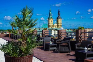 Terraço no telhado com móveis de vime, um vaso de planta e vista para um edifício histórico com duas torres.