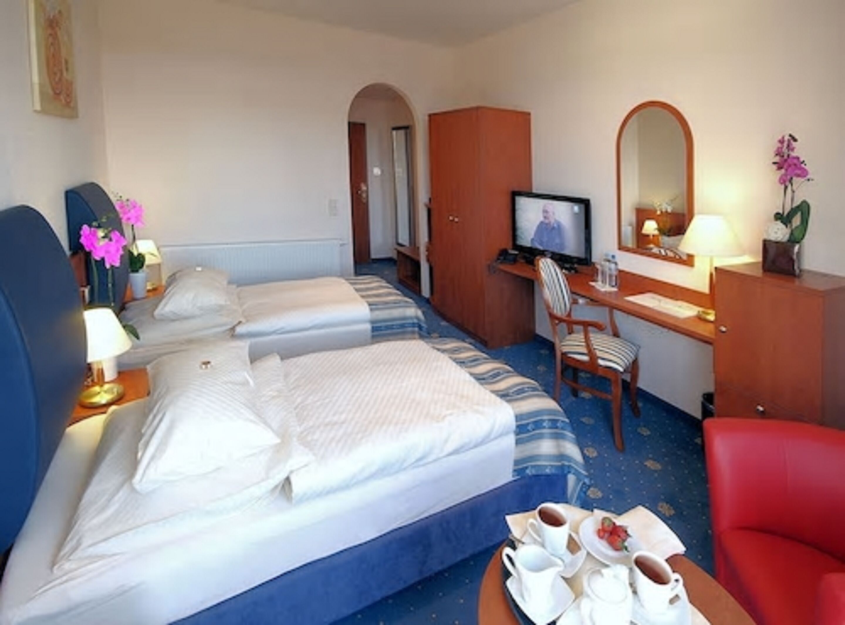Quarto de hotel com duas camas, uma escrivaninha com computador, televisão e uma cadeira vermelha.