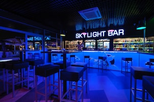 Um bar com uma placa de neon que diz "SKYLIGHT BAR" e um bartender. Há vários bancos e um balcão com uma variedade de garrafas e copos.