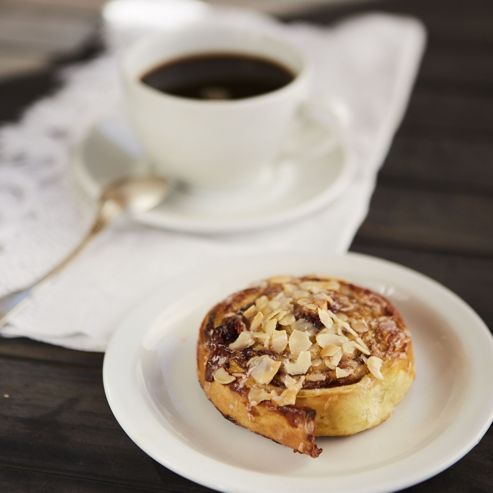 Um cinnamon roll sobre um prato branco, com uma xícara de café ao fundo.