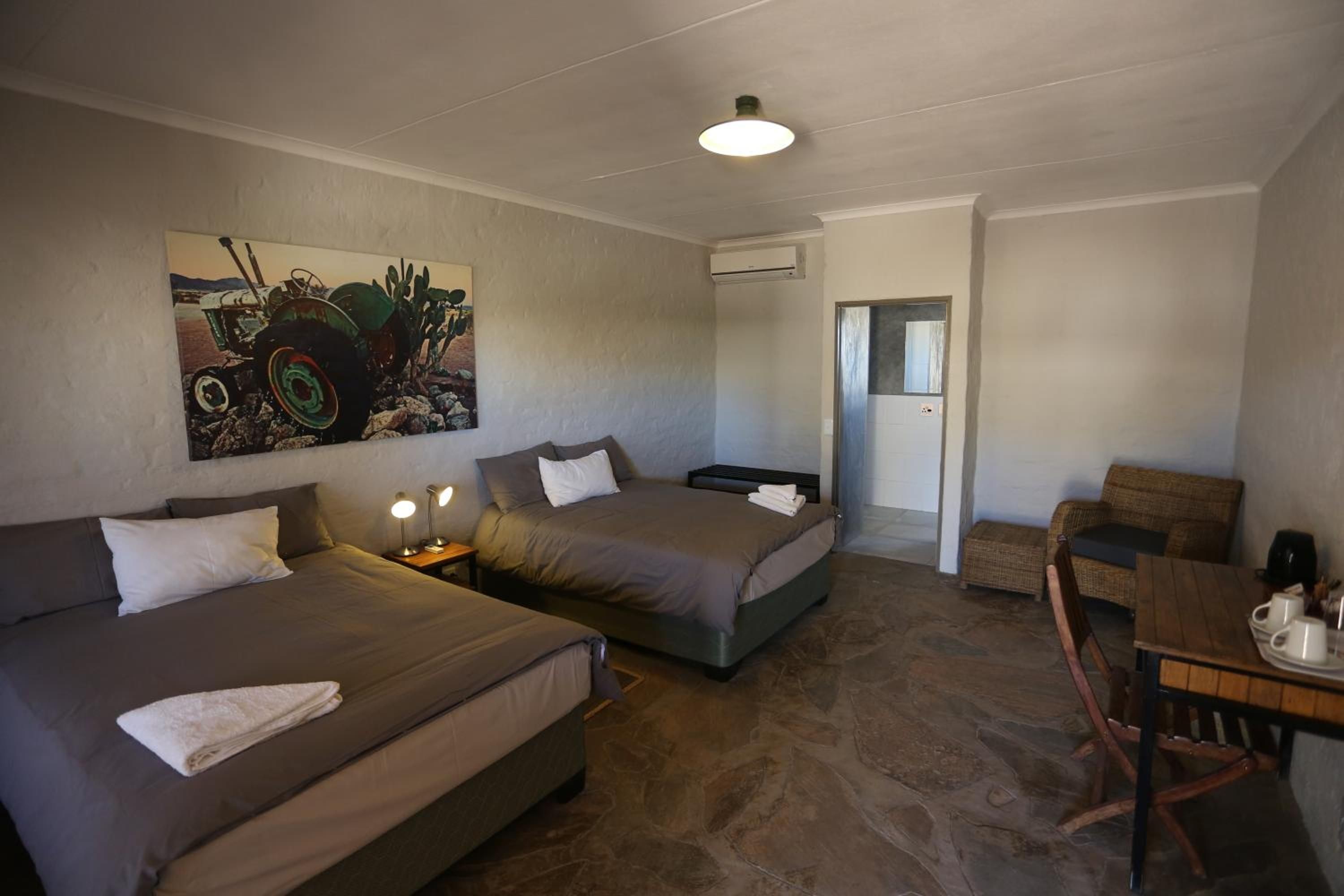 Quarto de hotel com duas camas, um quadro, uma escrivaninha e cadeiras, e um aparelho de ar condicionado.